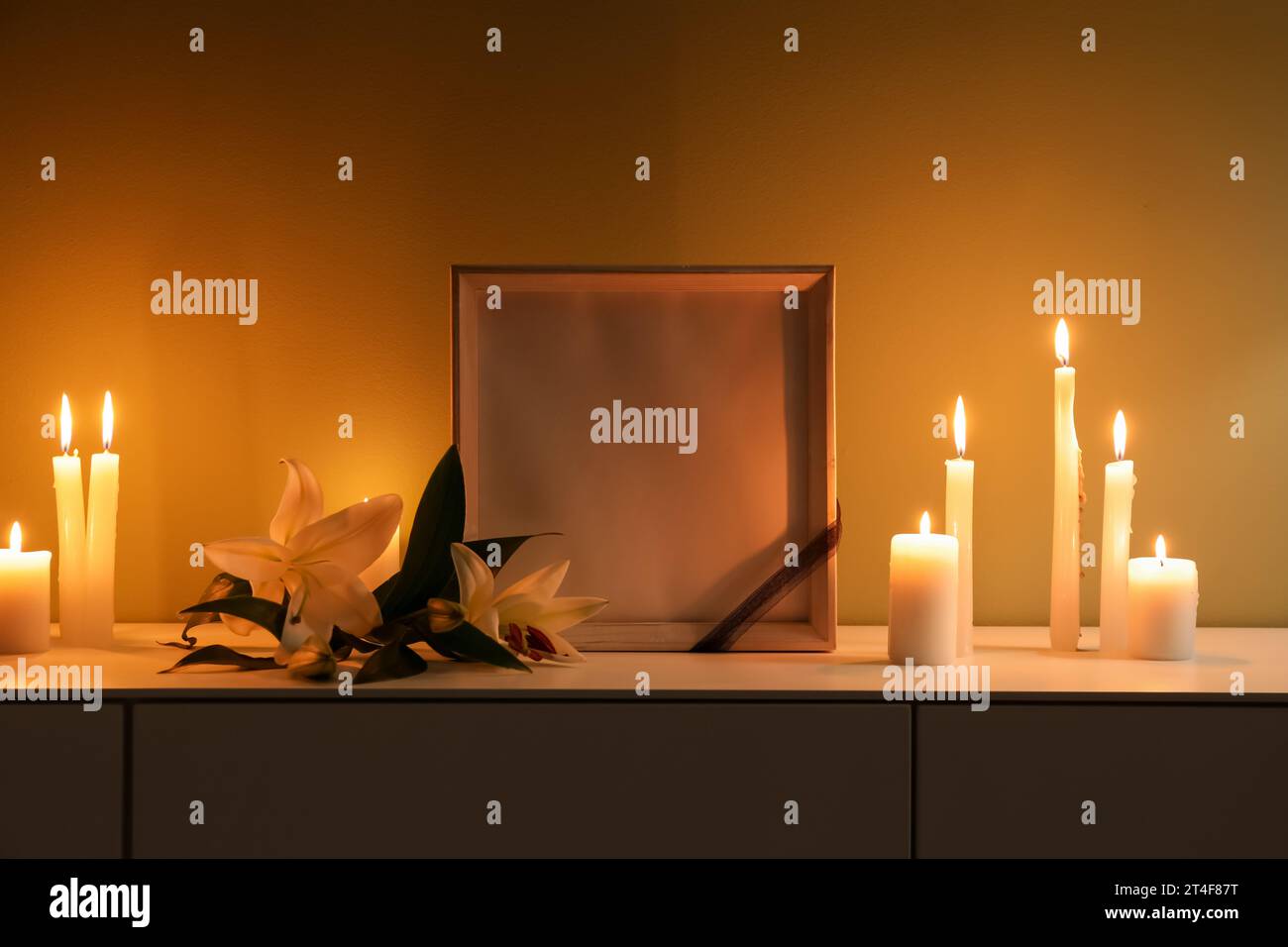 Blank funeral frame, burning candles and lily flowers on cabinet in ...