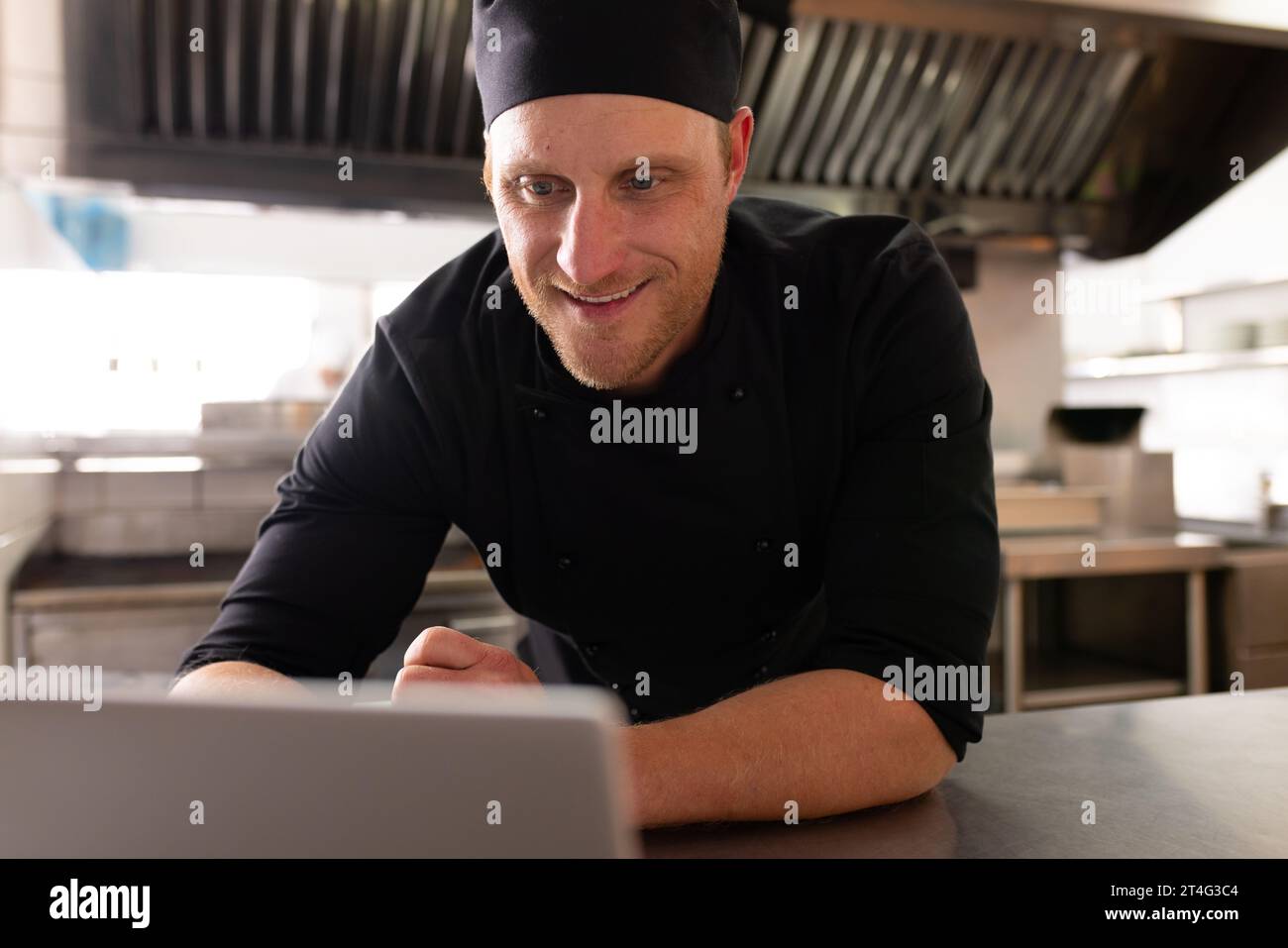 Smiling caucasian male chef in black uniform using laptop on counter in ...