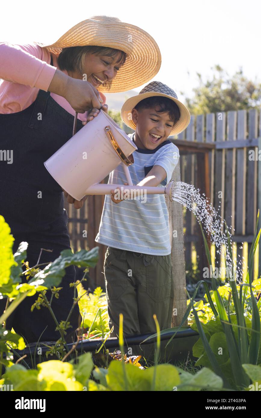 Happy Biracial Grandmother And Grandson Watering Plants In Sunny Garden
