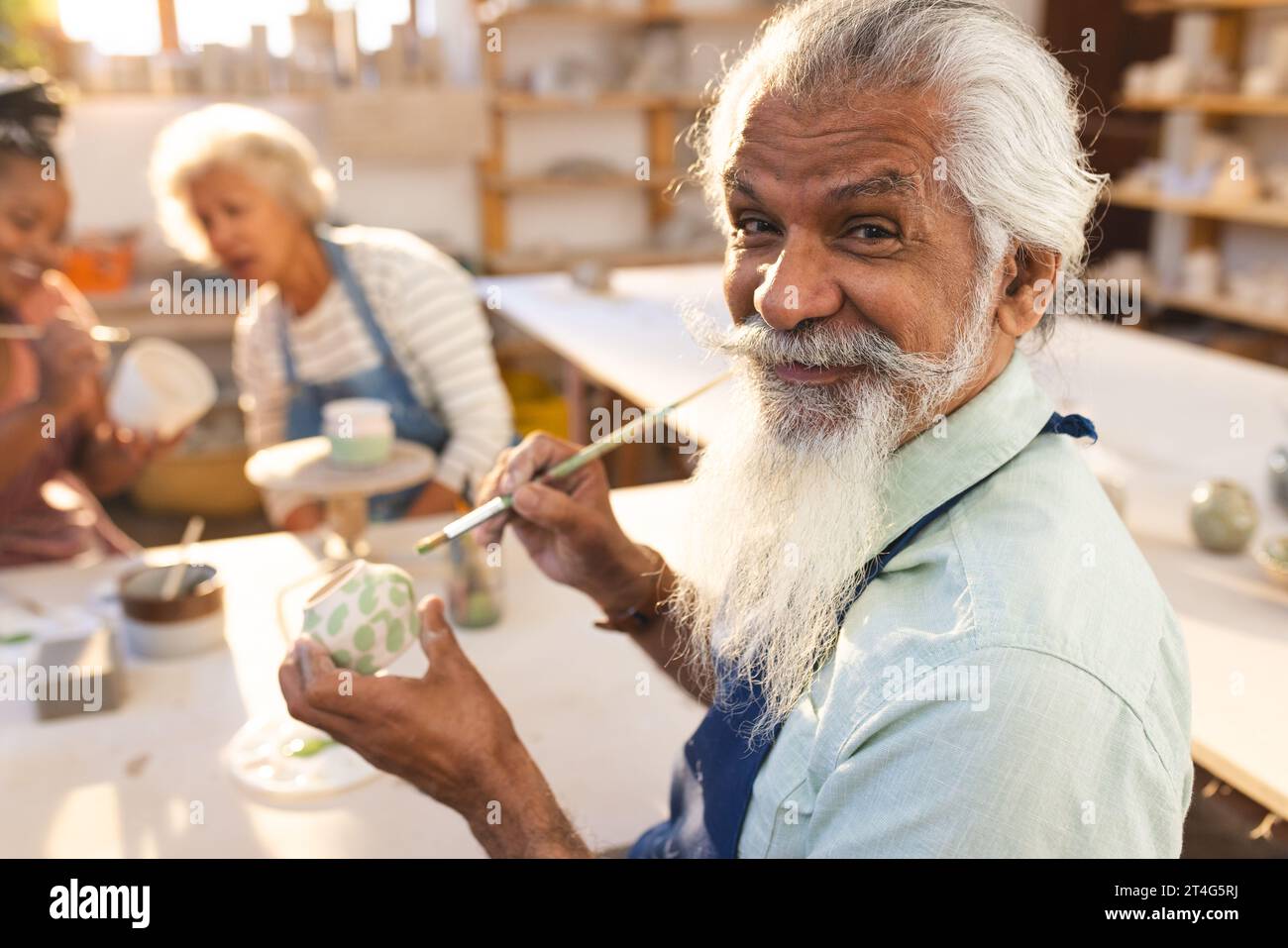 Happy biracial senior potter with long beard glazing clay jug and ...