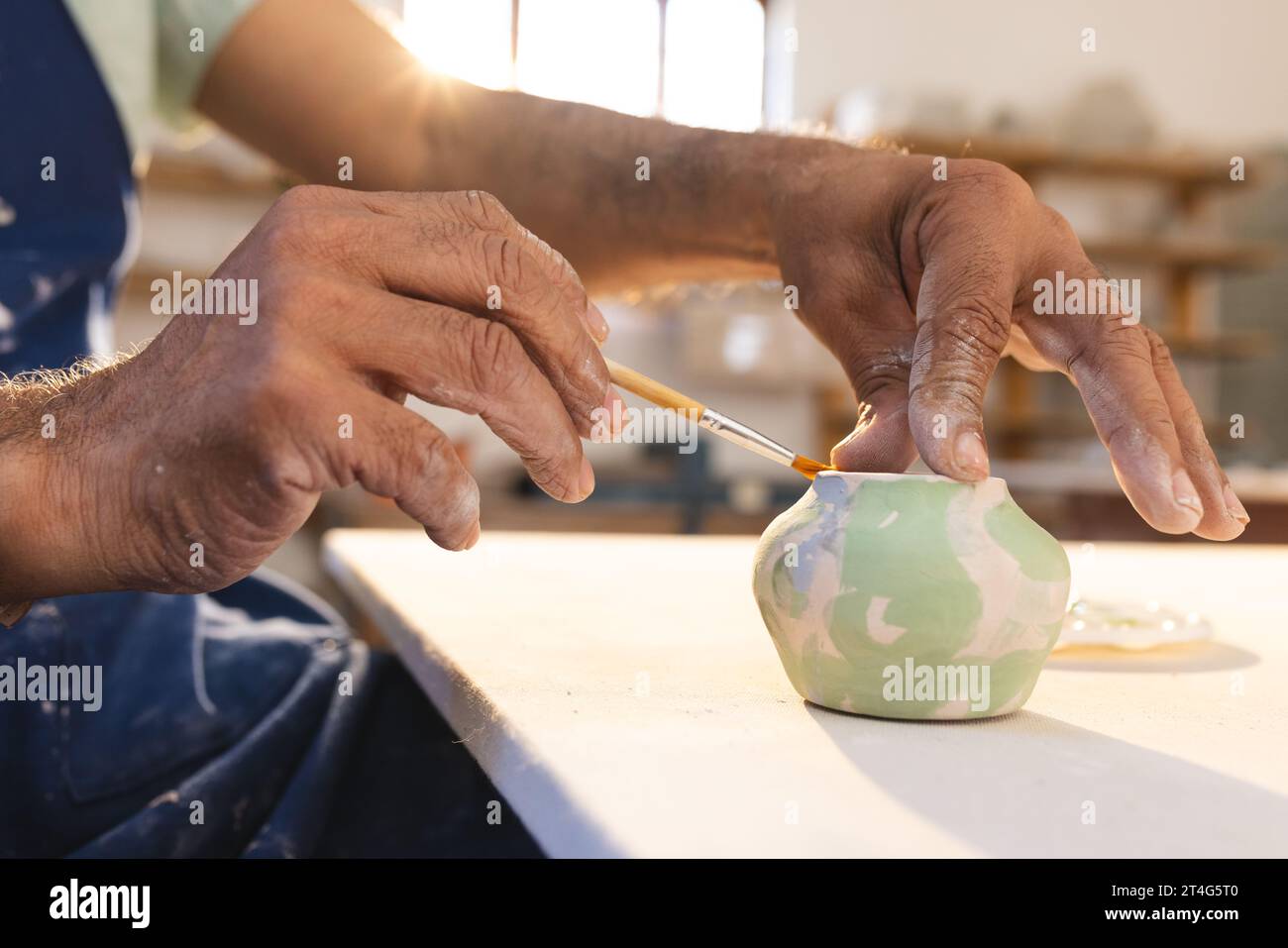 Hands of biracial senior male potter with glazing clay jug in pottery ...