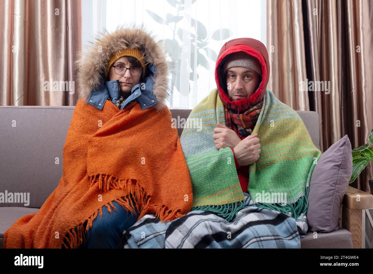 Sad middle age couple in warm clothes getting cold on the sofa in their ...
