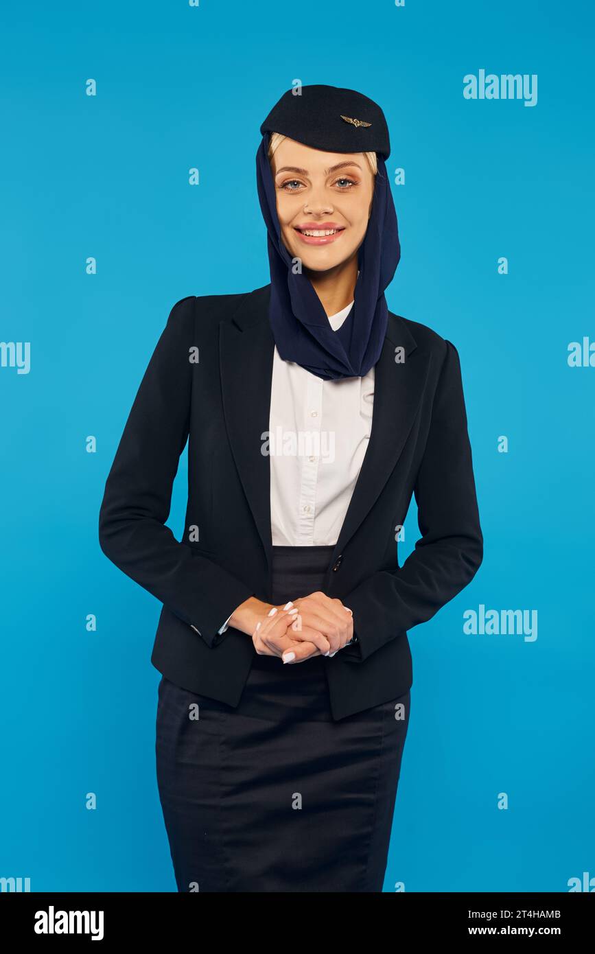 happy stewardess in uniform and headscarf smiling at camera on blue ...