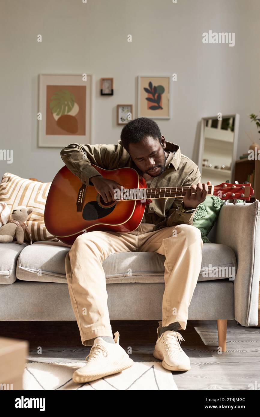 Vertical full length portrait of adult Black man playing guitar at home ...