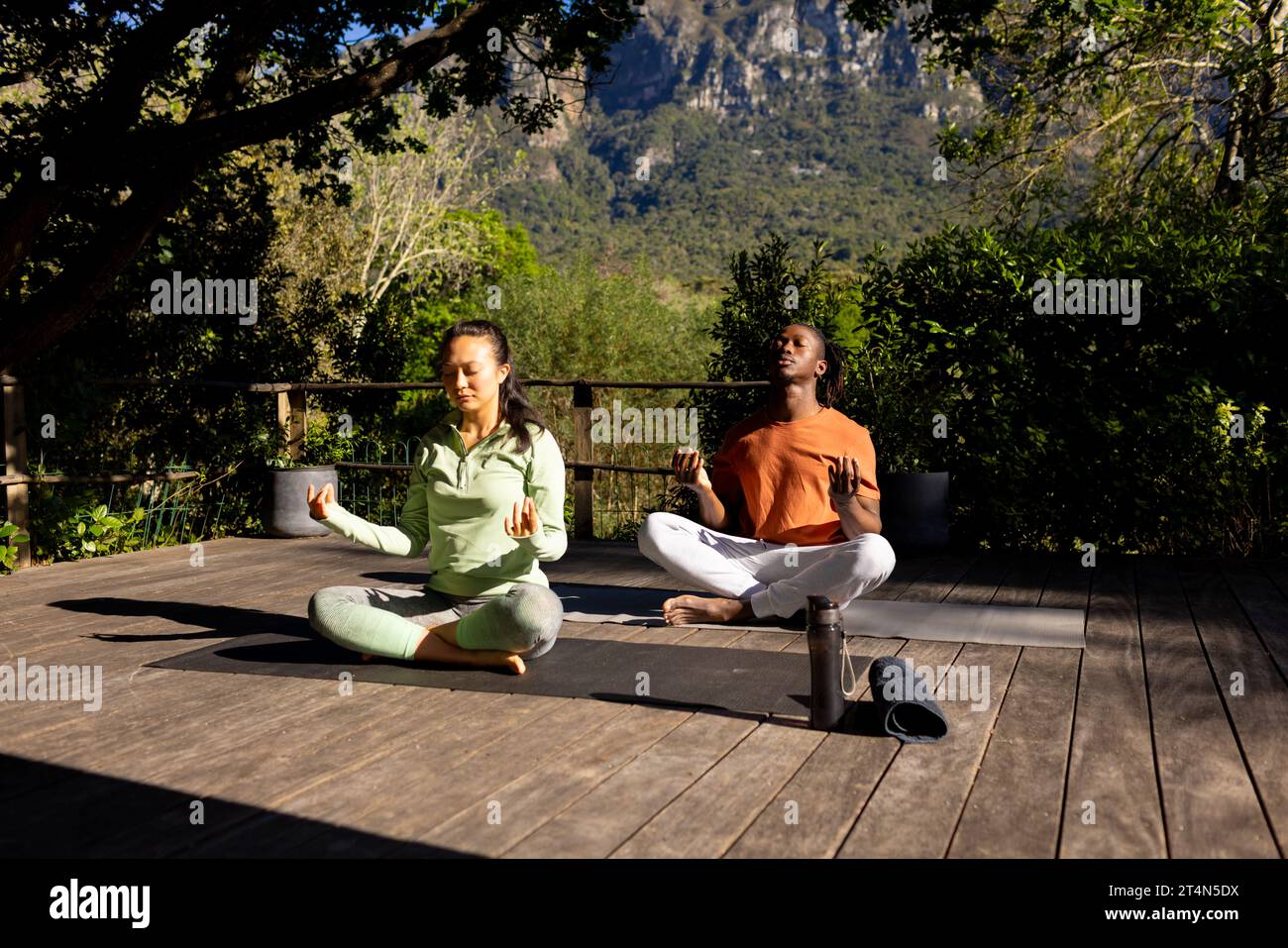 Happy diverse couple practicing yoga meditation sitting on deck in sunny garden, copy space ...