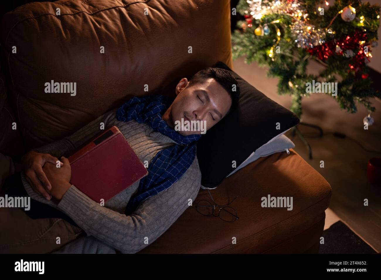 A tired Asian man falls asleep while reading a book, sleeping on a sofa ...