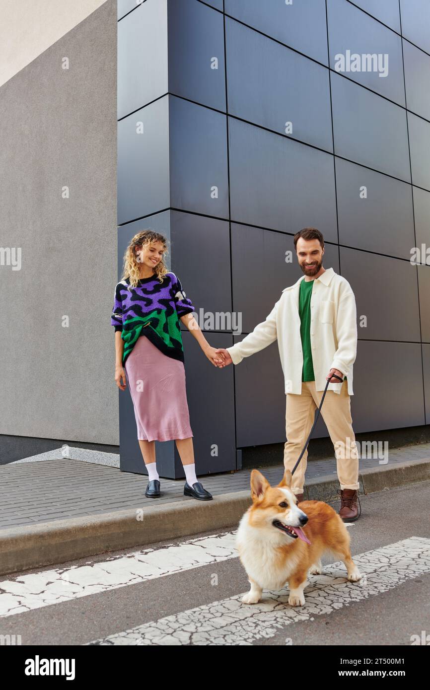 joyful couple holding hands and walking with corgi dog near modern grey ...