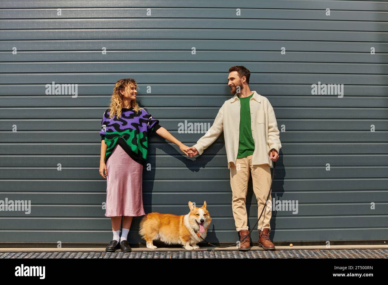 happy and stylish couple holding hands and standing with corgi dog near ...