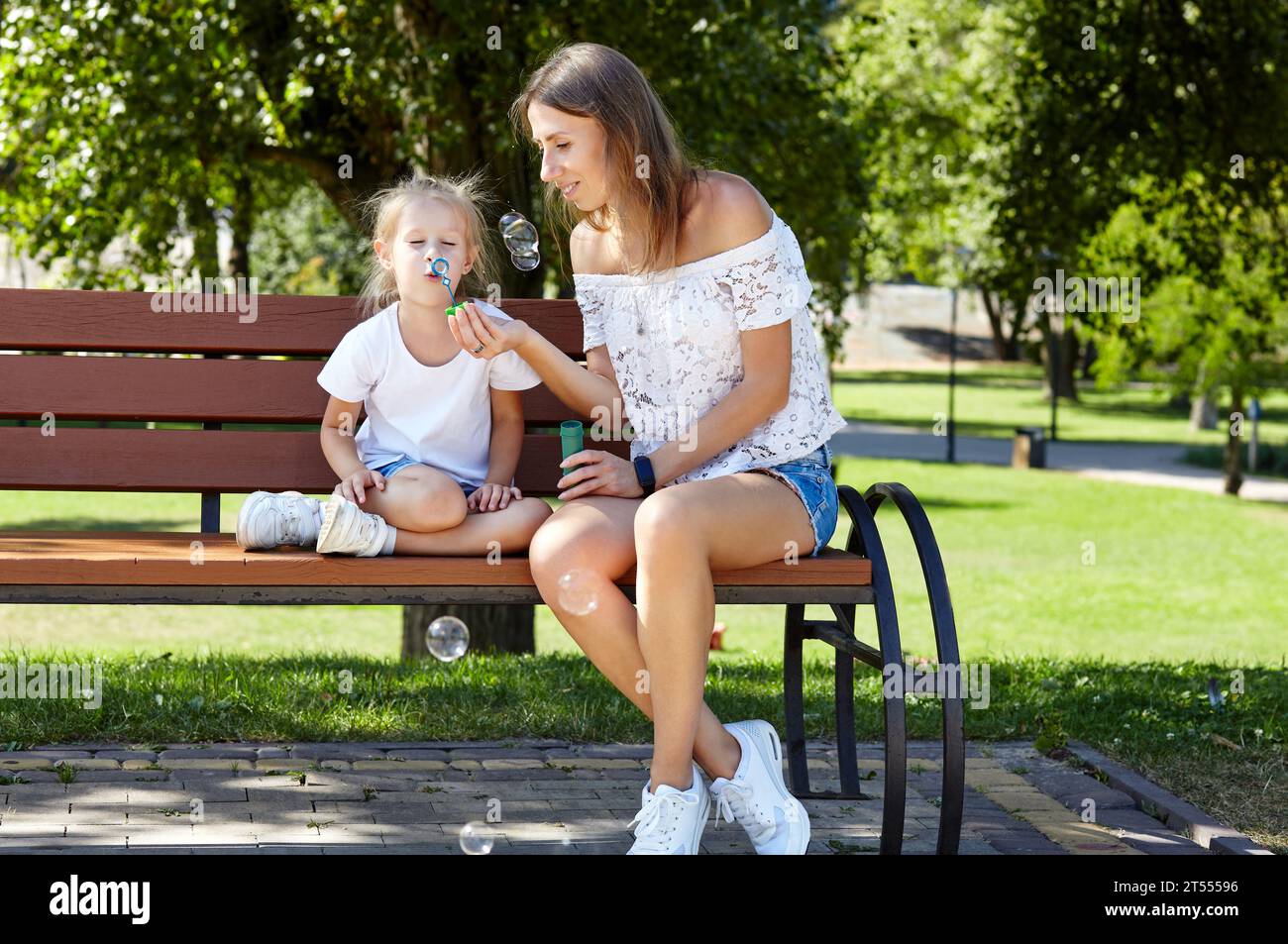 Mother and daughter sitting on a bench in the summer city park and blowing soap bubbles ...