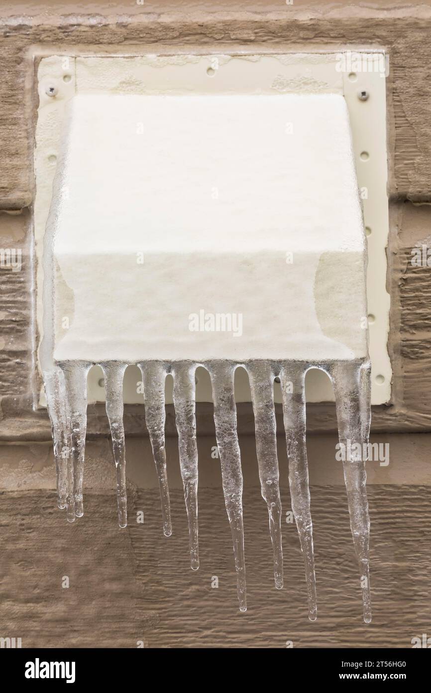 Metal exhaust vent covered with ice and icicles on tan clapboard wall ...
