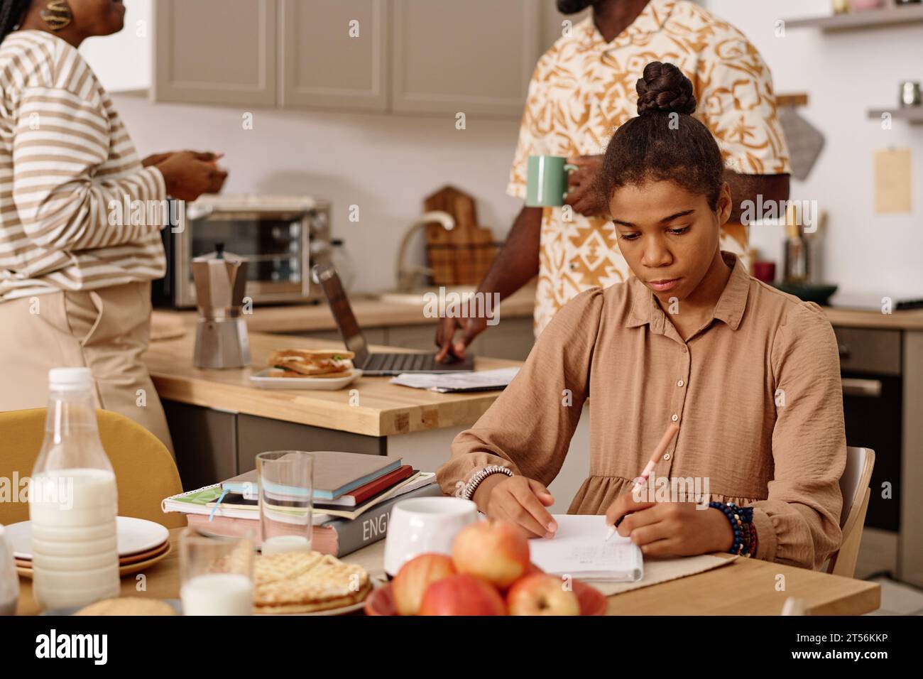 Serious teenage girl doing homework when parents eating breakfast in ...