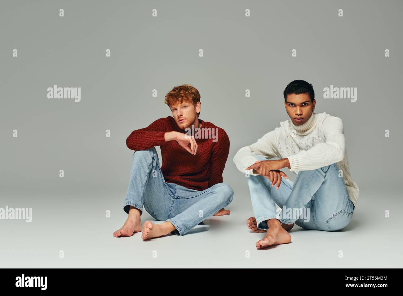 multicultural handsome male models in warm sweaters sitting on floor looking at camera, men ...