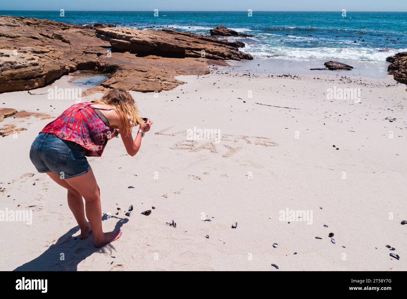 A woman in denim shorts takes a picture of writing in the sand at ...