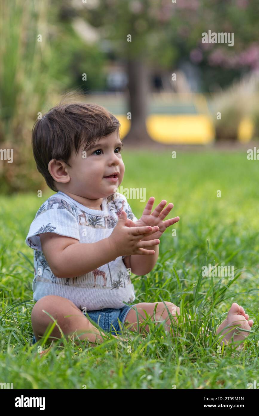 Cute baby clapping and playing while sitting on the grass in a park ...