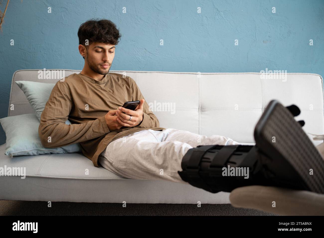 Man with leg injury lying on sofa with phone, ankle brace orthosis on male leg at home Stock ...