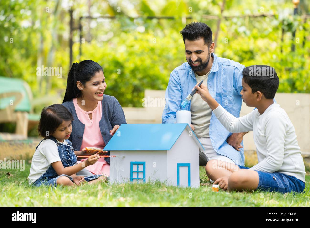 Happy indian parents helping kids to do painting on toy house at park ...