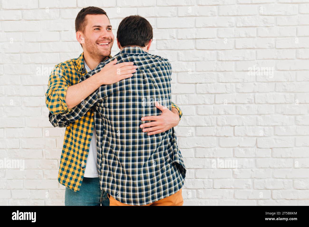 Smiling portrait man giving hug his friend against white brick wall ...