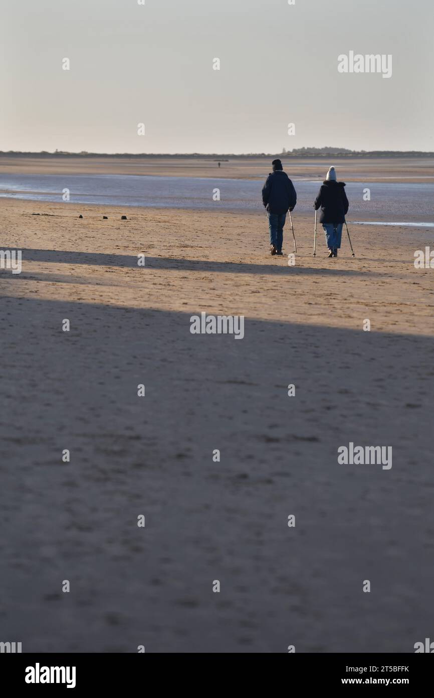 couple walking away from camera with sticks low tide brancaster beach ...