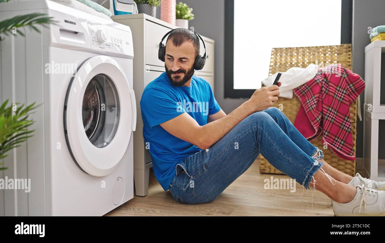 Young hispanic man listening to music waiting for washing machine at ...