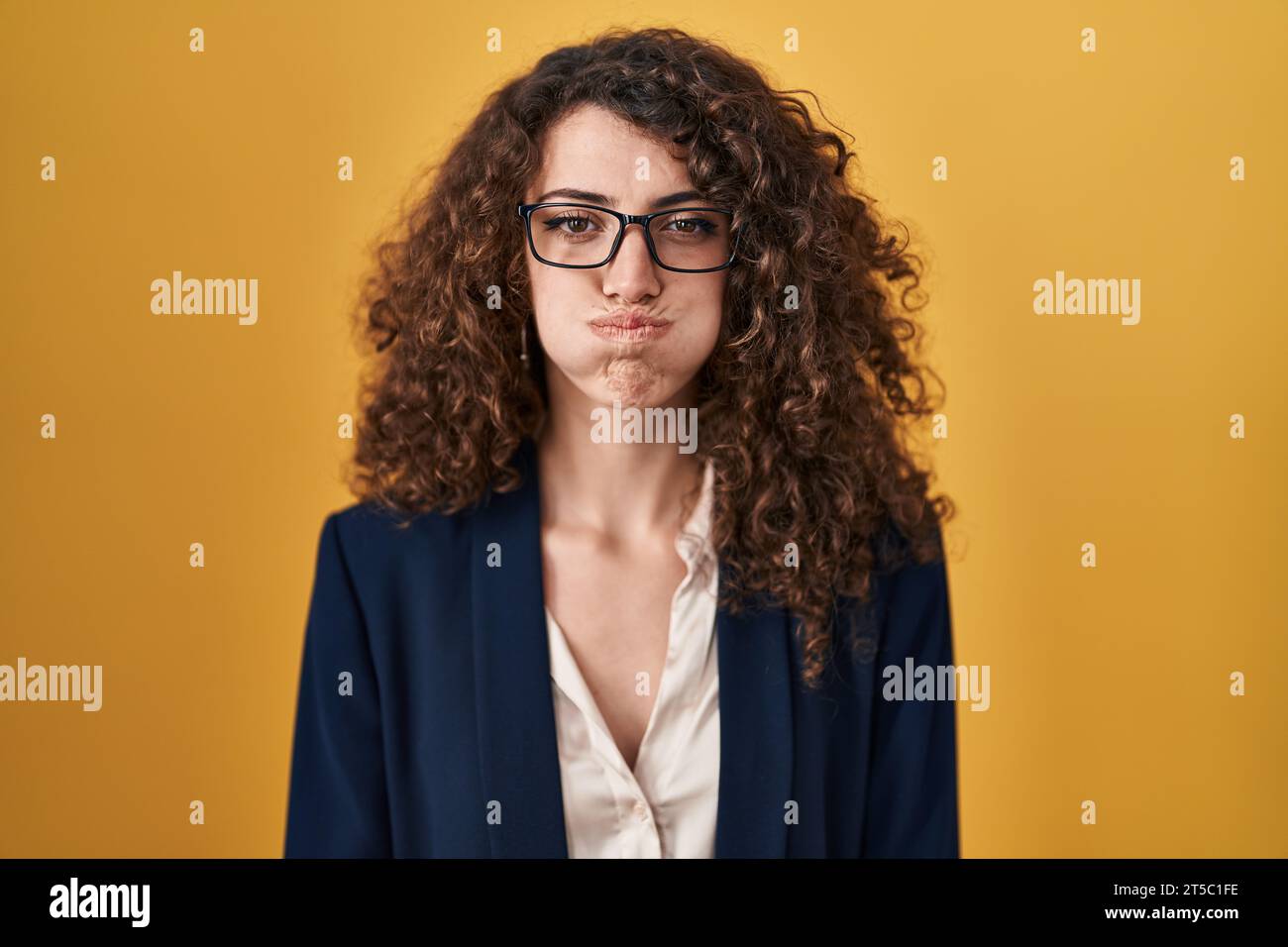 Hispanic woman with curly hair standing over yellow background puffing ...