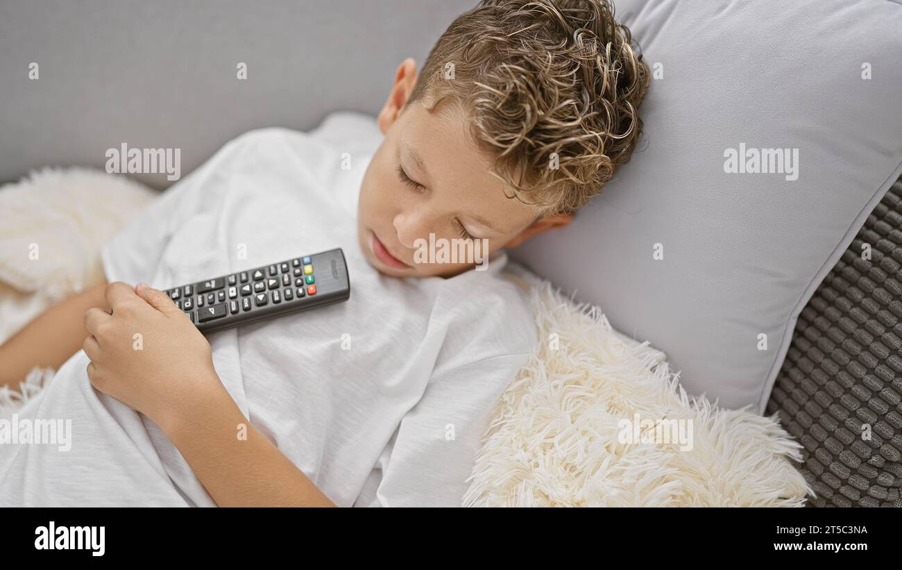 Blond boy holding tv remote control lying on sofa sleeping at home ...