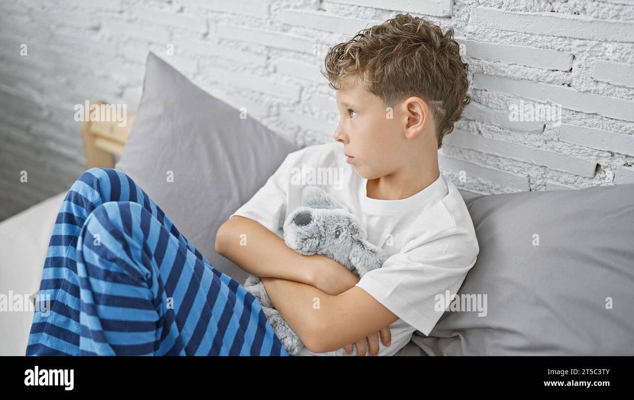 Adorable blond boy in pyjamas, sitting on bed in his bedroom, hugging ...