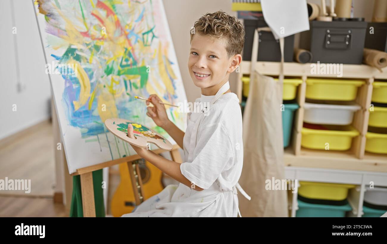 Adorable blond boy artist, confidently smiling as he enjoys drawing at ...