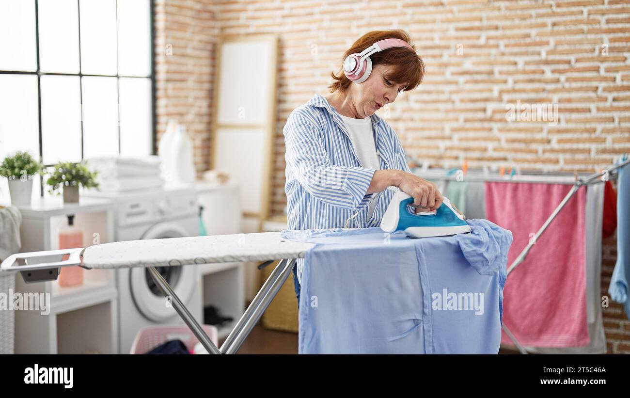 Middle age woman listening to music ironing clothes at laundry room ...
