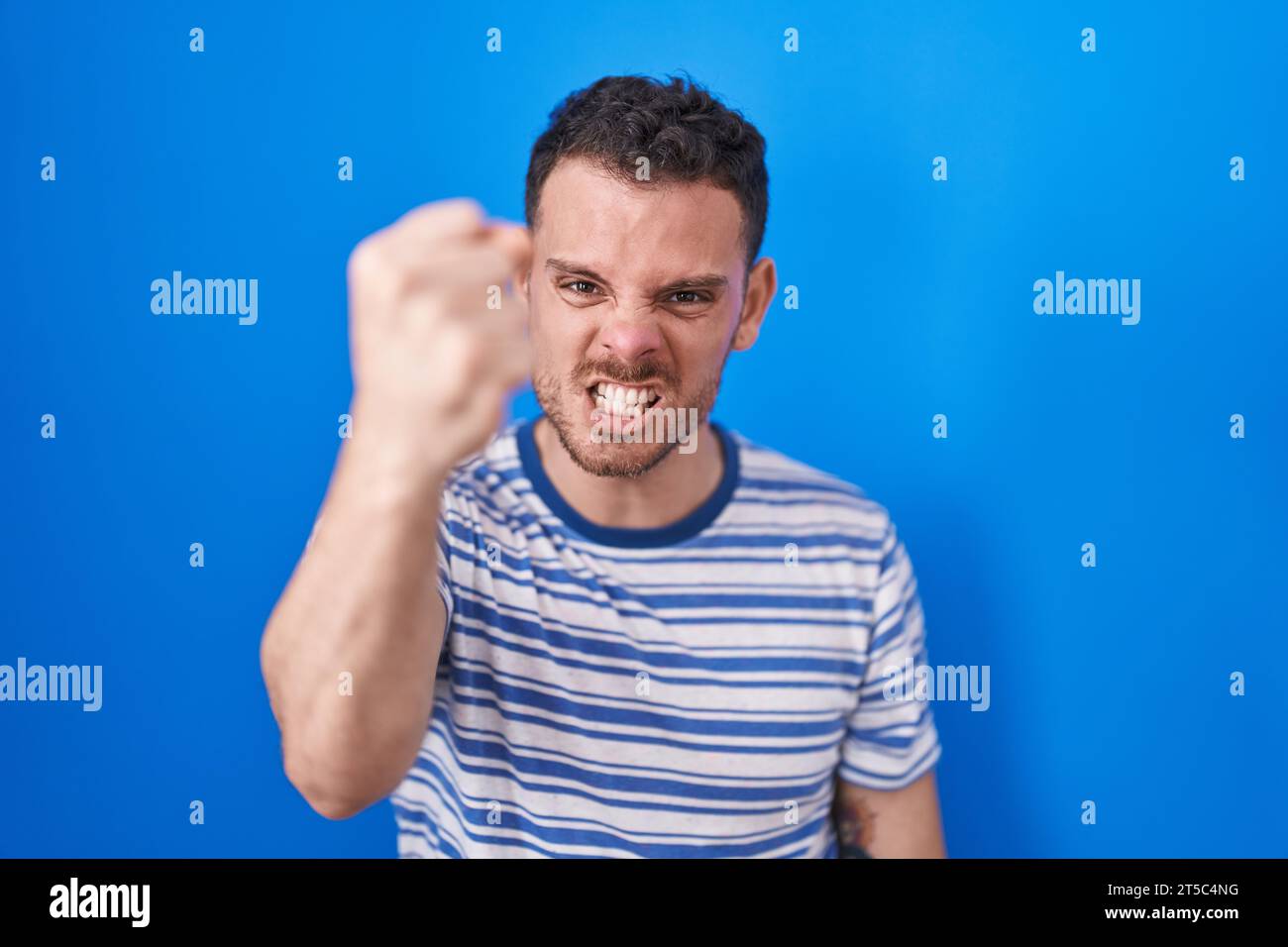 Young hispanic man standing over blue background angry and mad raising ...