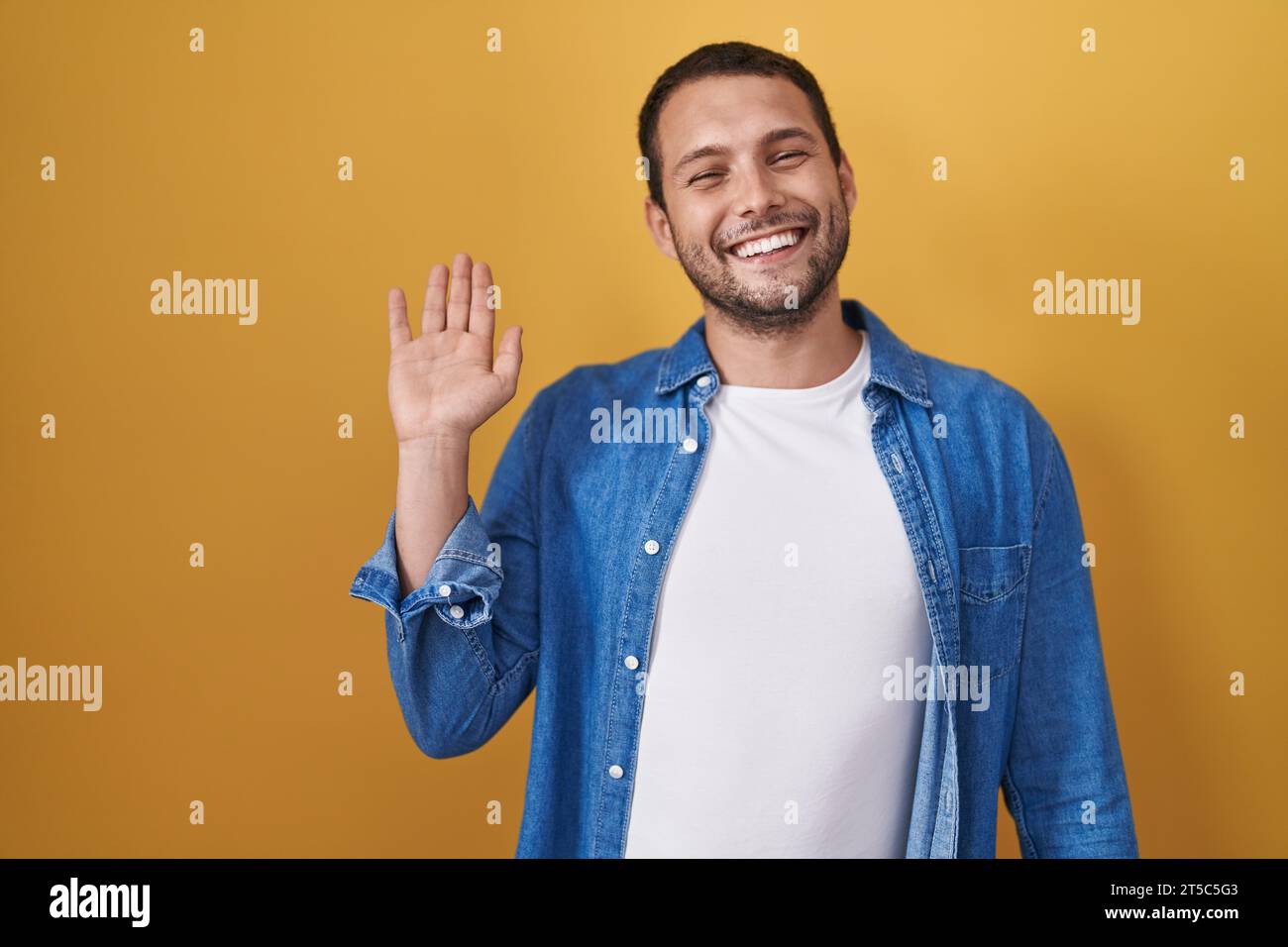 Hispanic man standing over yellow background waiving saying hello happy ...