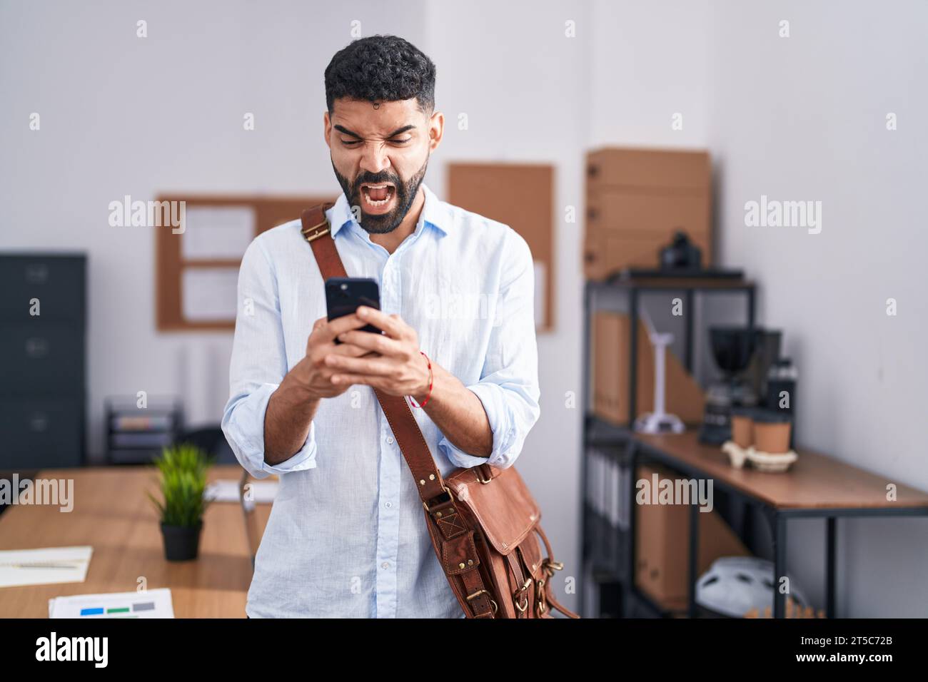Hispanic man with beard using smartphone at the office angry and mad screaming frustrated and ...
