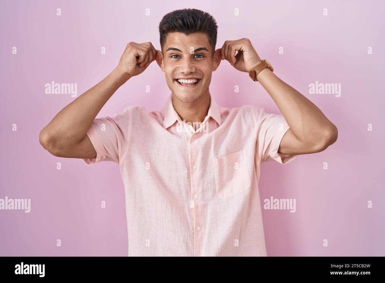 Young hispanic man standing over pink background smiling pulling ears ...