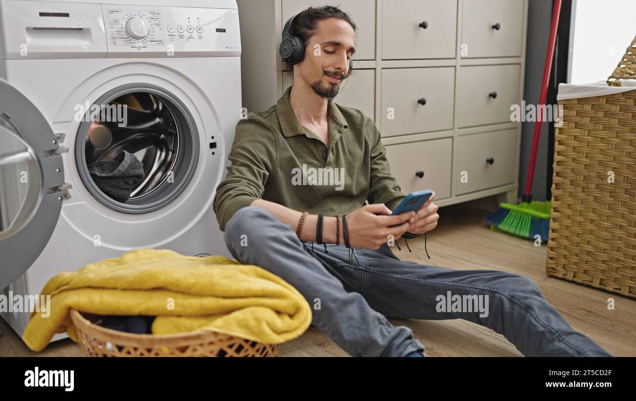 Young hispanic man listening to music leaning on washing machine at ...