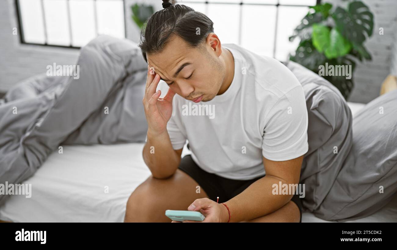 Stressed young chinese man, sitting in bedroom, using smartphone. under ...