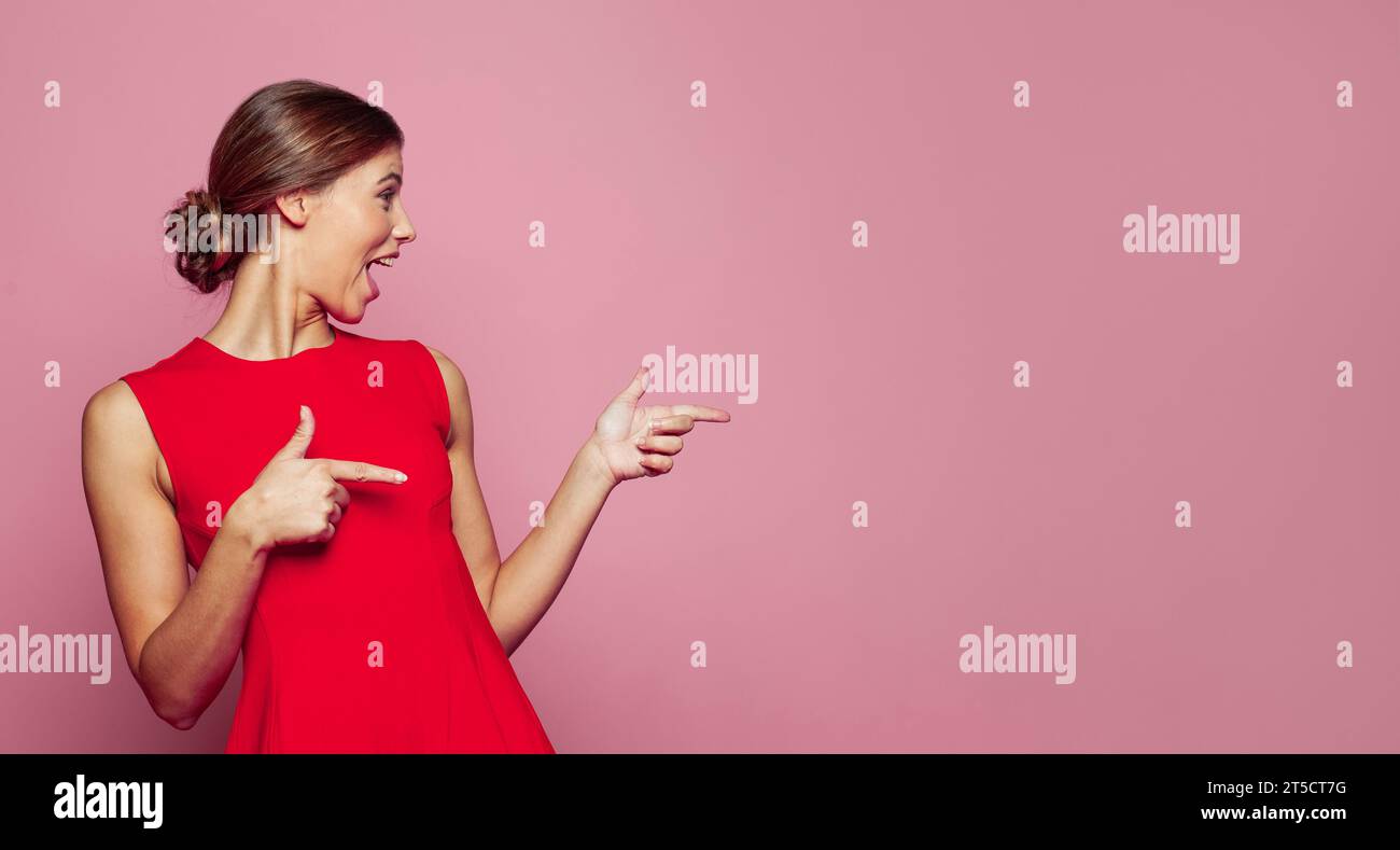 Happy beautiful woman pointing finger against pink studio wall banner ...