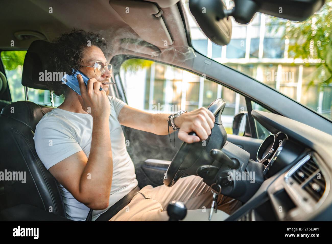 Man with curly hair driving a car, smoking a cigarette and using phone ...