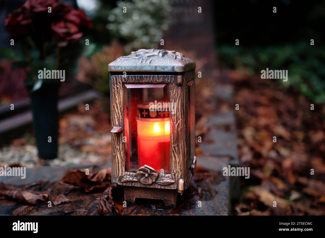 Ornate metal grave lantern with a burning candle on a grave with autumn leaves on All Saints ...