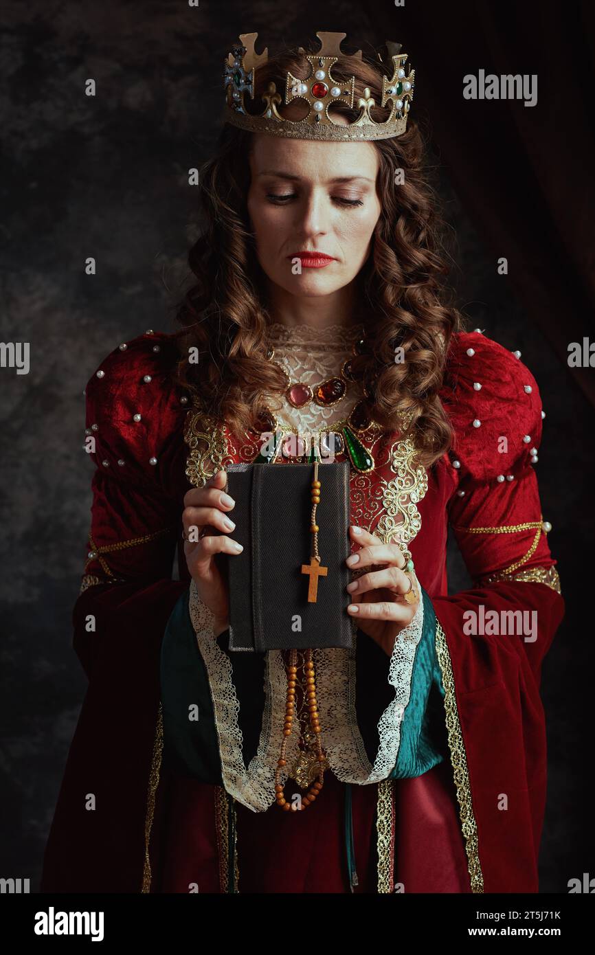 medieval queen in red dress with book, rosary and crown on dark gray ...