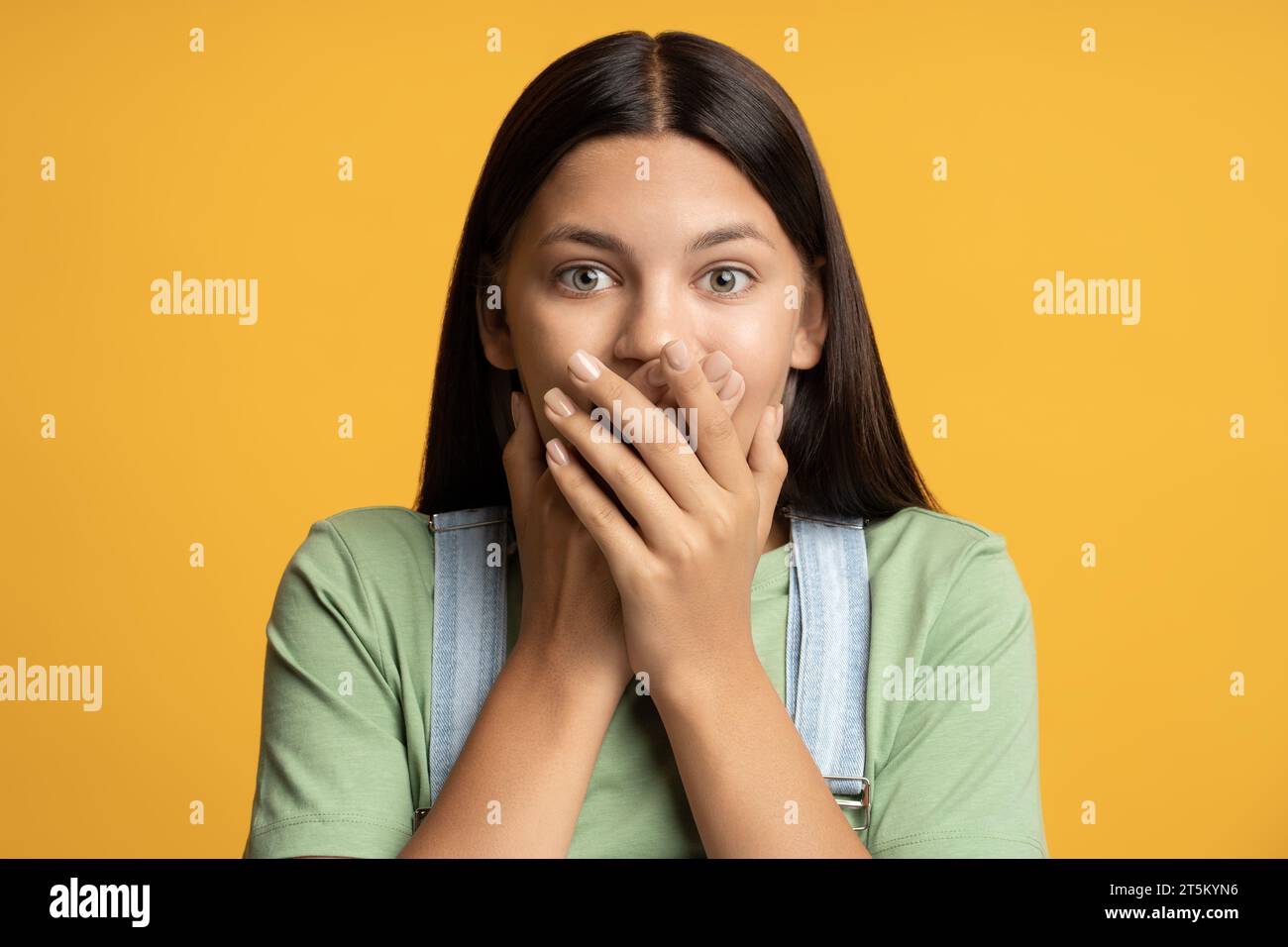 Stressed teenager with big round eyes closing open mouth with hands in ...