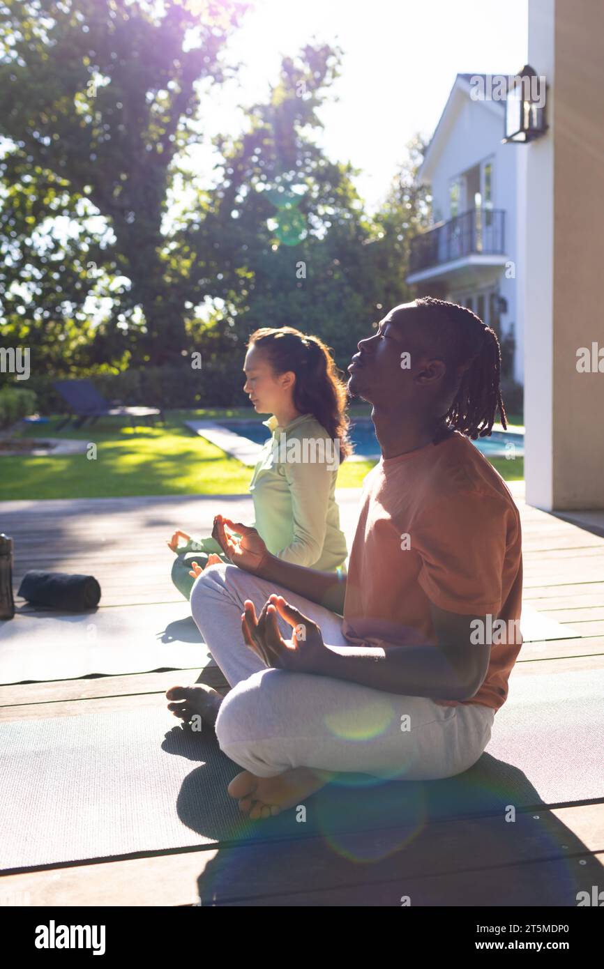 Happy diverse couple practicing yoga meditation sitting on deck in sunny garden, copy space ...