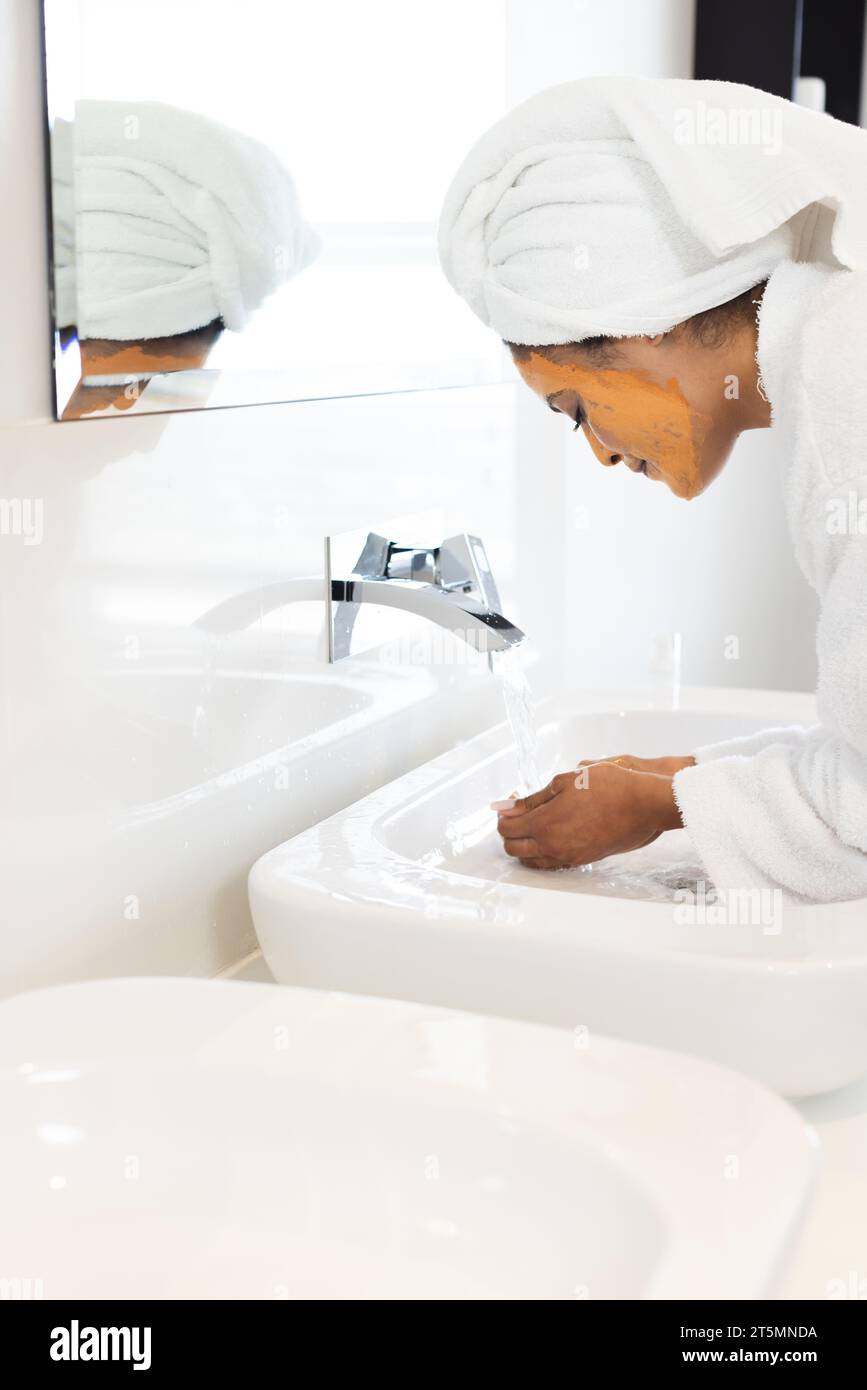 Happy biracial woman wearing bathrobe and washing her face in front of ...