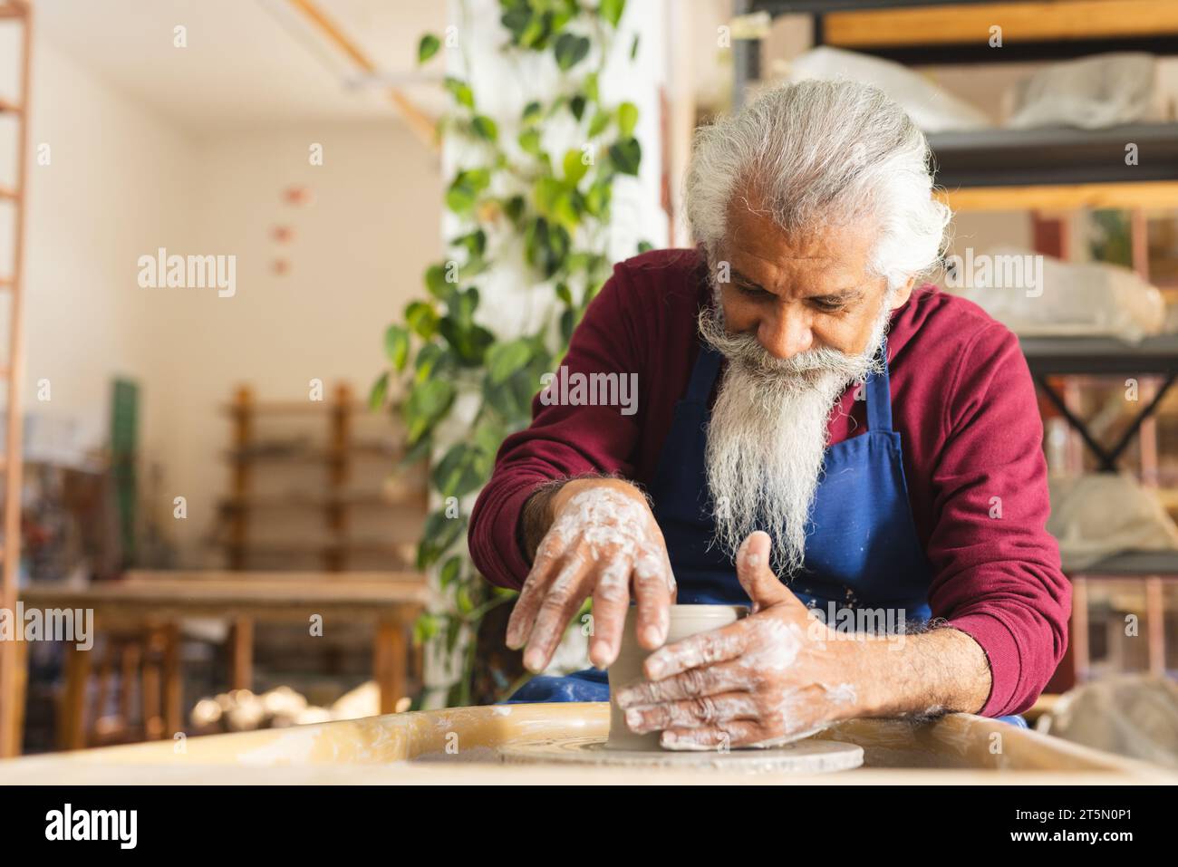 Focused biracial senior potter with long beard using potter's wheel in ...
