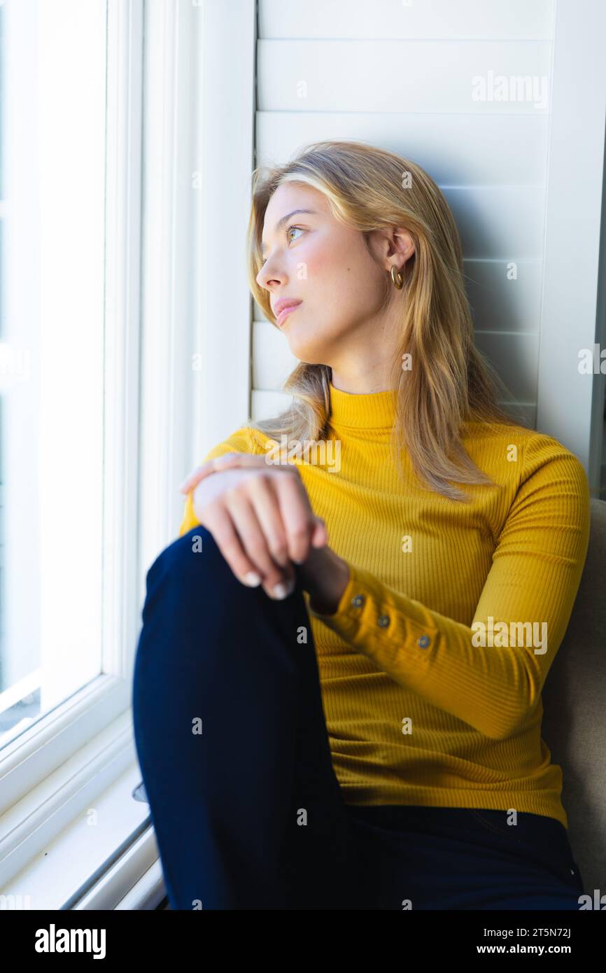 Pensive caucasian woman sitting on windowsill and looking out window in ...
