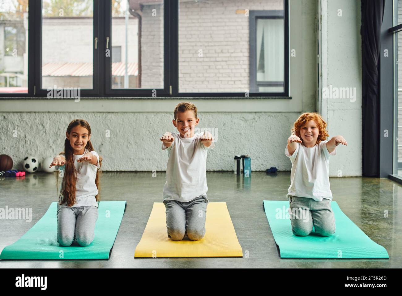 little joyful children sitting on fitness mats and stretching arms ...