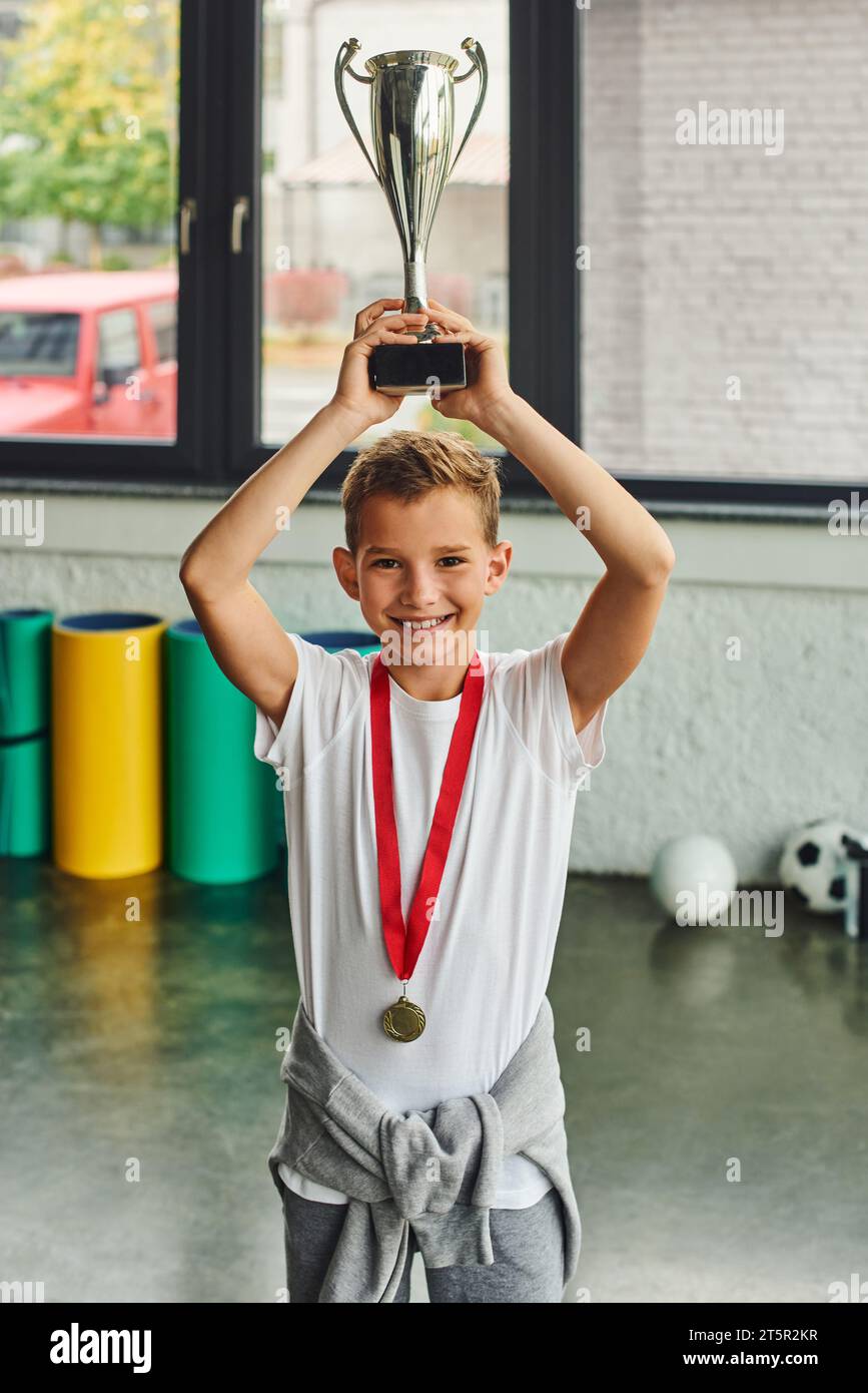 vertical shot of cheerful little boy with golden medal raising trophy ...