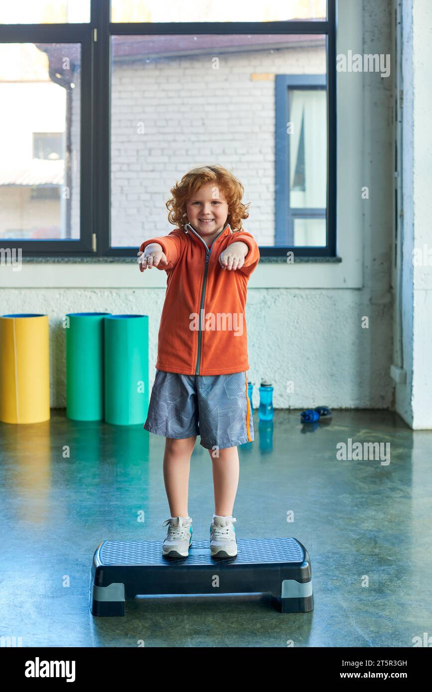 vertical shot of jolly little boy stretching arms on fitness stepper ...