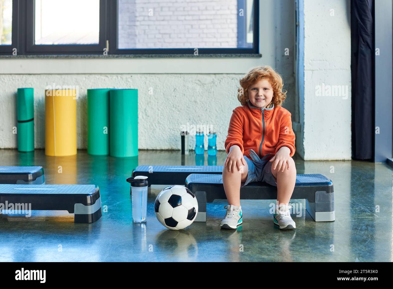 joyful red haired boy sitting on fitness stepper next to soccer ball ...