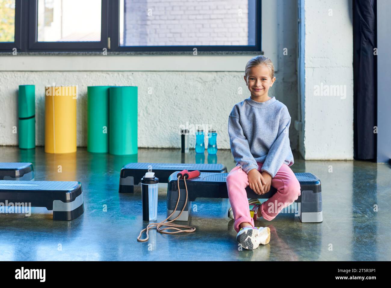 cheerful blonde little girl sitting on fitness stepper in sportswear ...