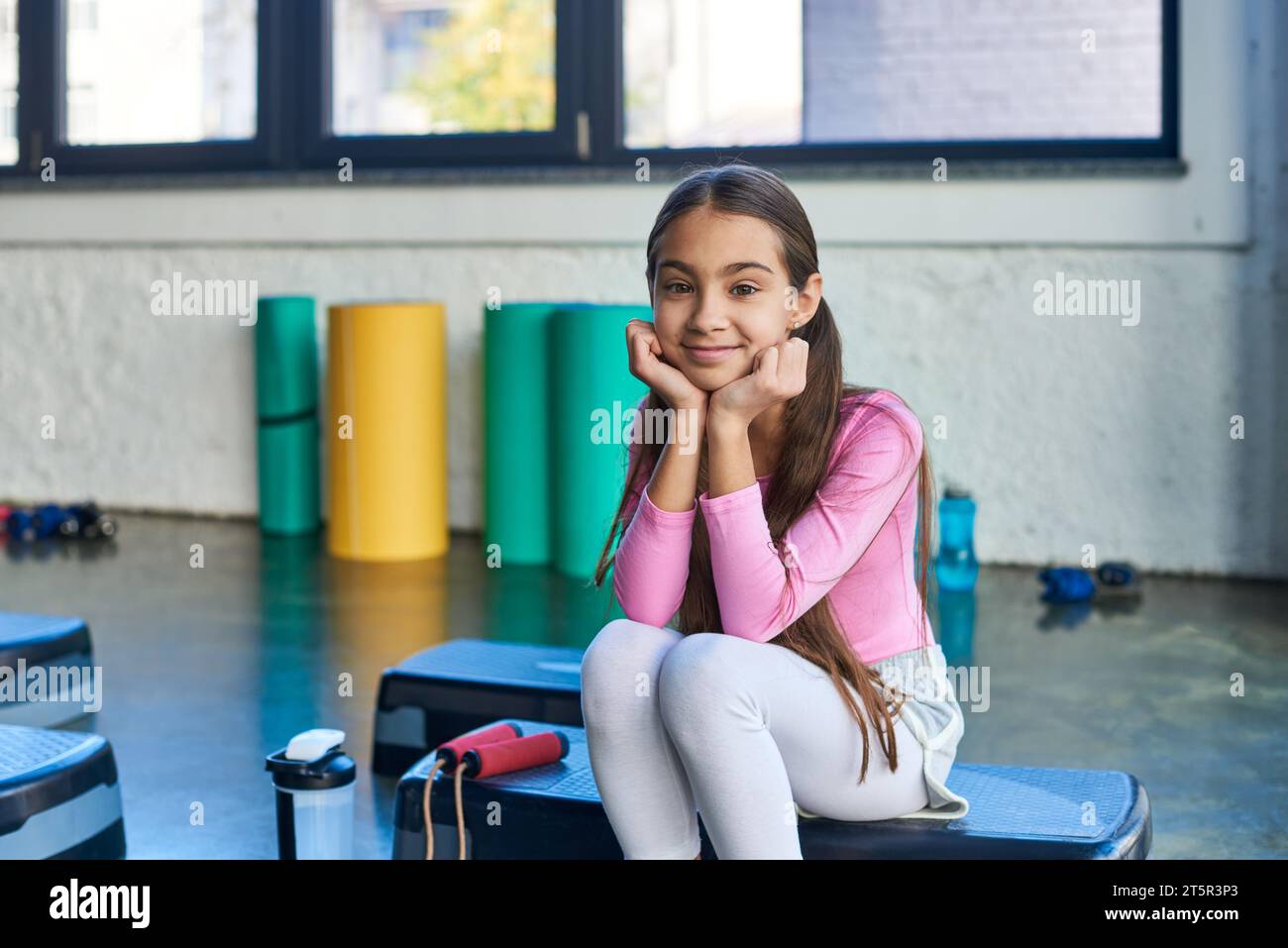 joyful girl sitting on fitness stepper with jump rope smiling at camera ...