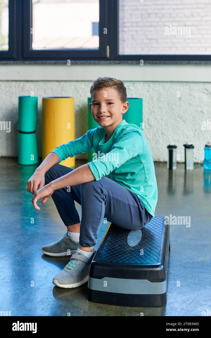 joyful boy in sportswear sitting on fitness stepper and smiling happily ...